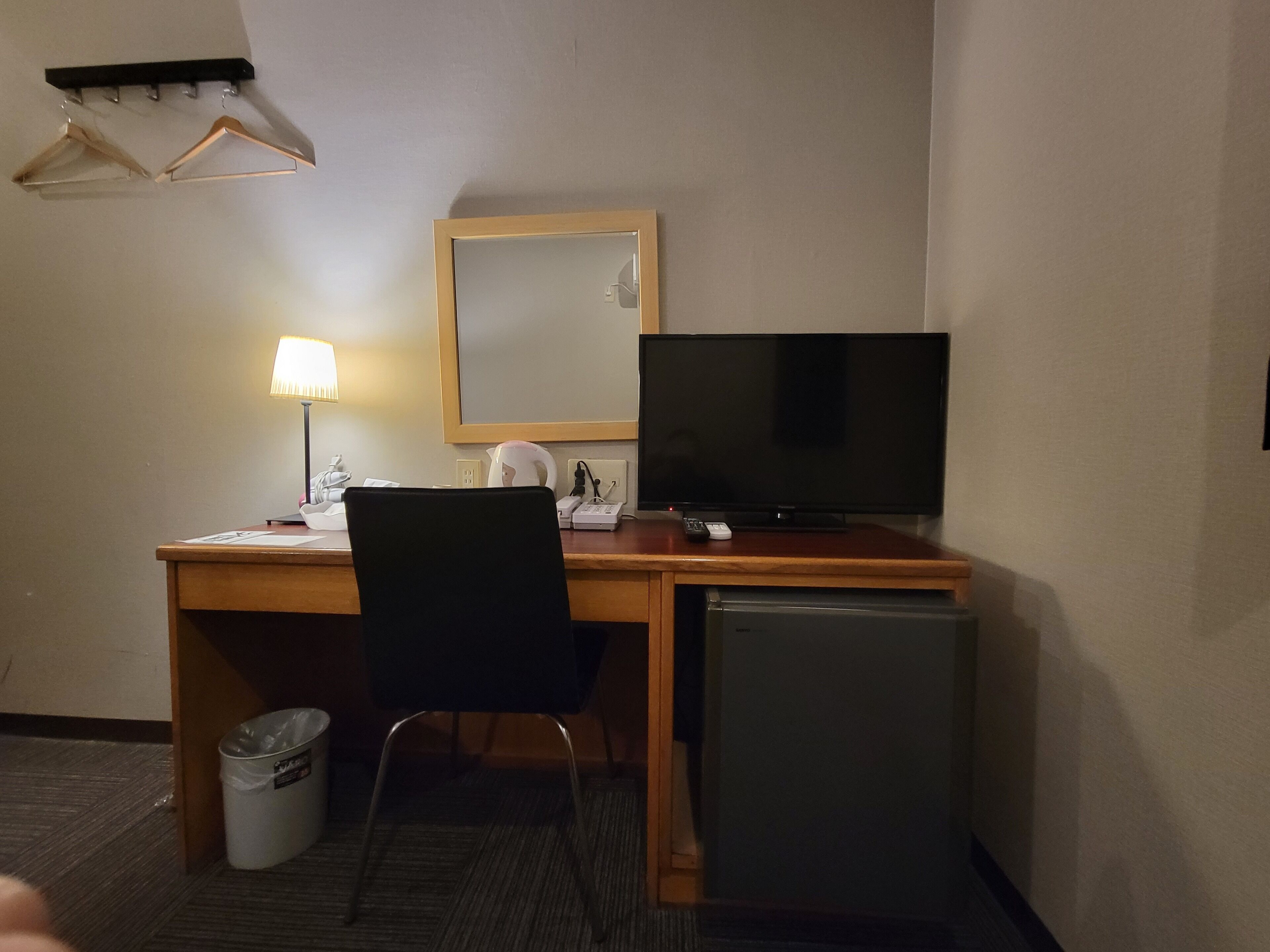 Quarto de hotel com escrivaninha de madeira, uma cadeira preta, televisão, mini-geladeira, lixeira e luminária.