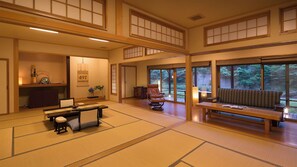 Um quarto tradicional japonês com tatames, uma mesa baixa com almofada, uma cadeira estofada e um sofá com vista para o exterior.
