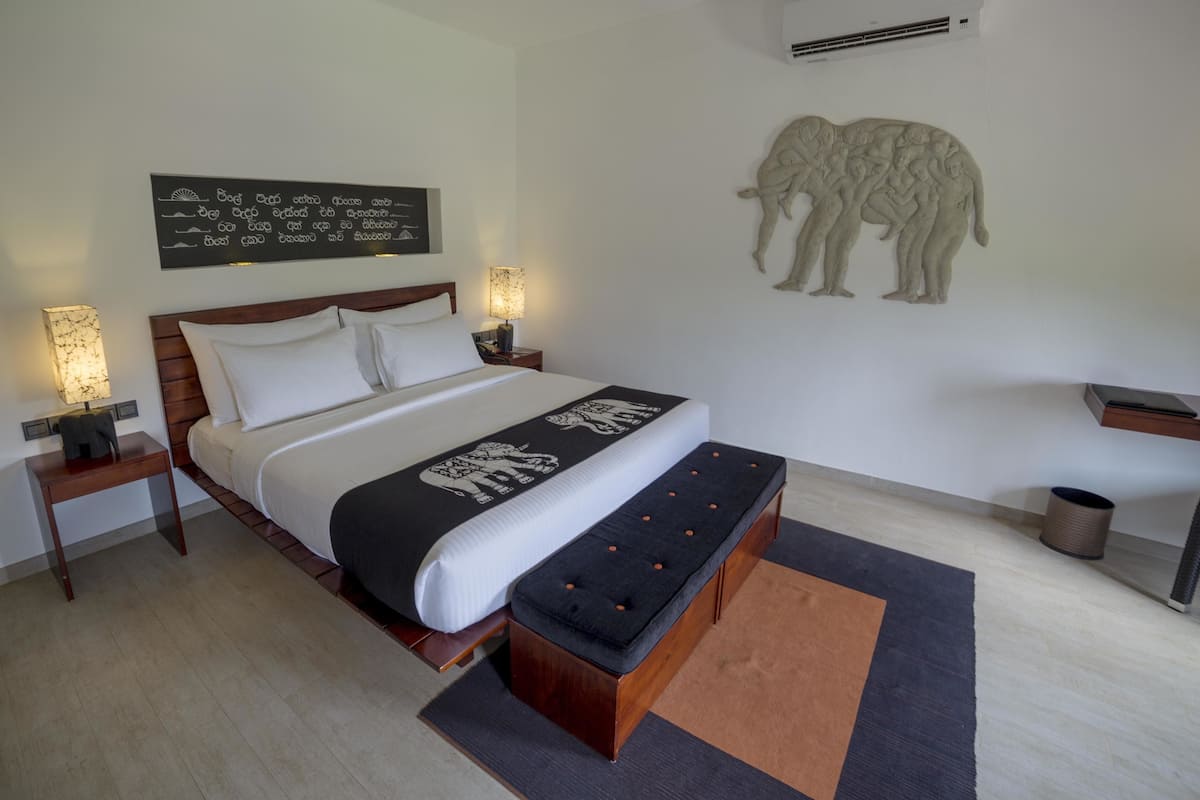 Quarto bem arrumado com cama grande, mesas de cabeceira com luminárias, ar condicionado na parede e uma escultura decorativa de elefante.
