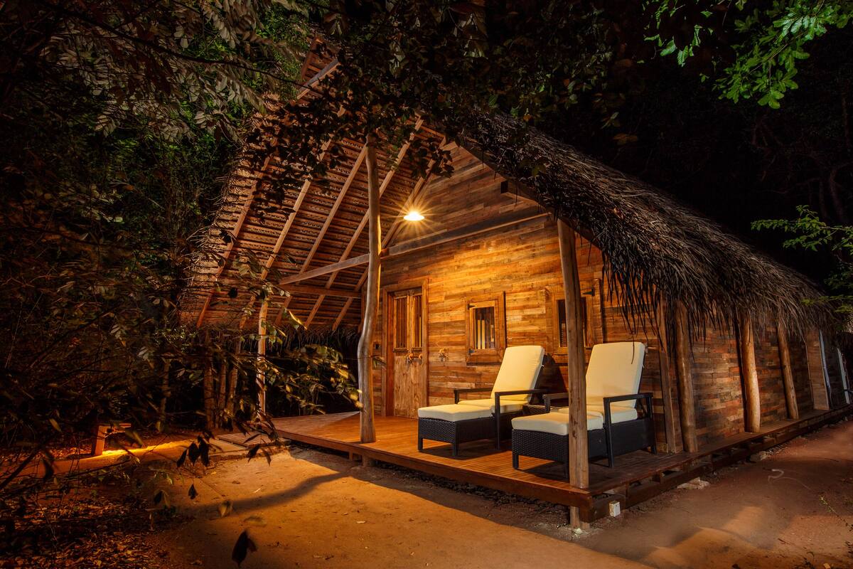 Uma cabana de madeira com telhado de palha, duas poltronas de descanso e um alpendre.