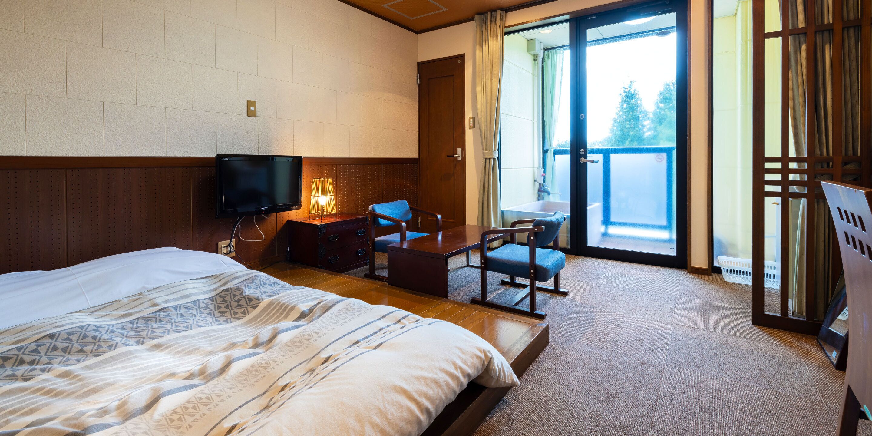 Quarto de hotel com uma cama grande, uma mesa com cadeira, televisão e varanda com vista para árvores.