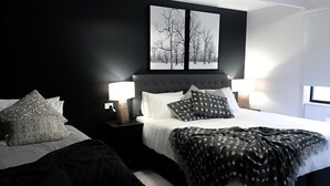 Quarto moderno com parede escura, cama grande com cabeceira estofada, duas mesas de cabeceira com abajures e uma janela que proporciona luz natural.