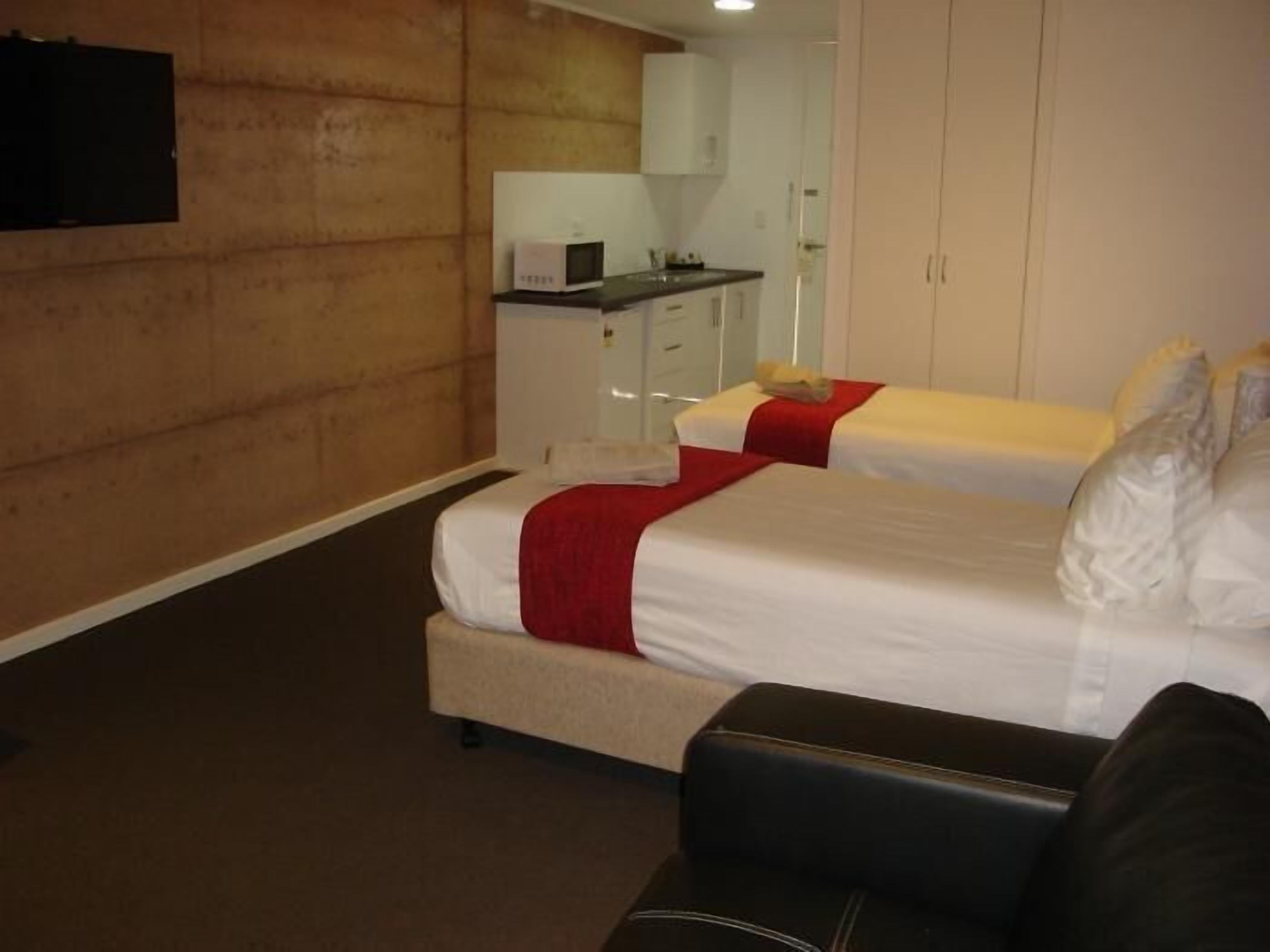 Quarto de hotel com duas camas, uma kitchenette e uma televisão fixada na parede.