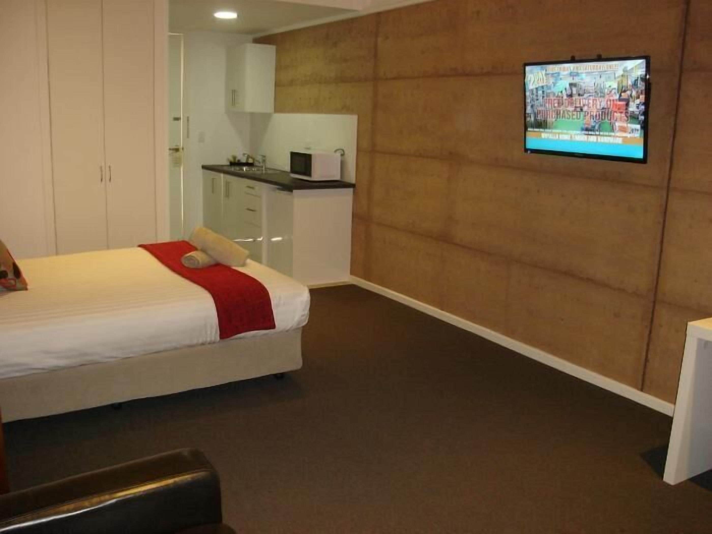Quarto de hotel com cama, TV fixada na parede, pequena copa e parede de destaque marrom.