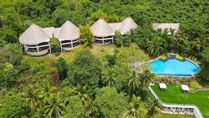 Vista aérea de um resort com villas de telhado de palha, piscina e vegetação tropical exuberante.