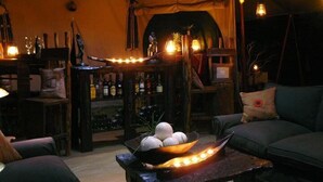 Um quarto aconchegante, com iluminação suave e decoração rústica, apresenta uma mesa de madeira com uma tigela de objetos esféricos brancos, um sofá com um travesseiro e uma prateleira com garrafas.