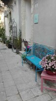 Uma viela estreita com um banco de metal azul e uma mesa rosa com flores.