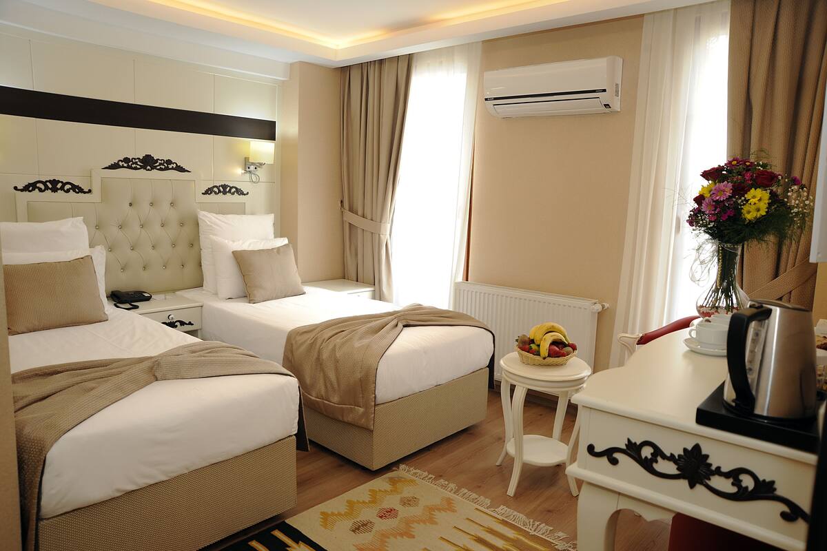 Quarto de hotel com duas camas, uma mesa pequena com uma fruteira, chaleira e um vaso com flores.