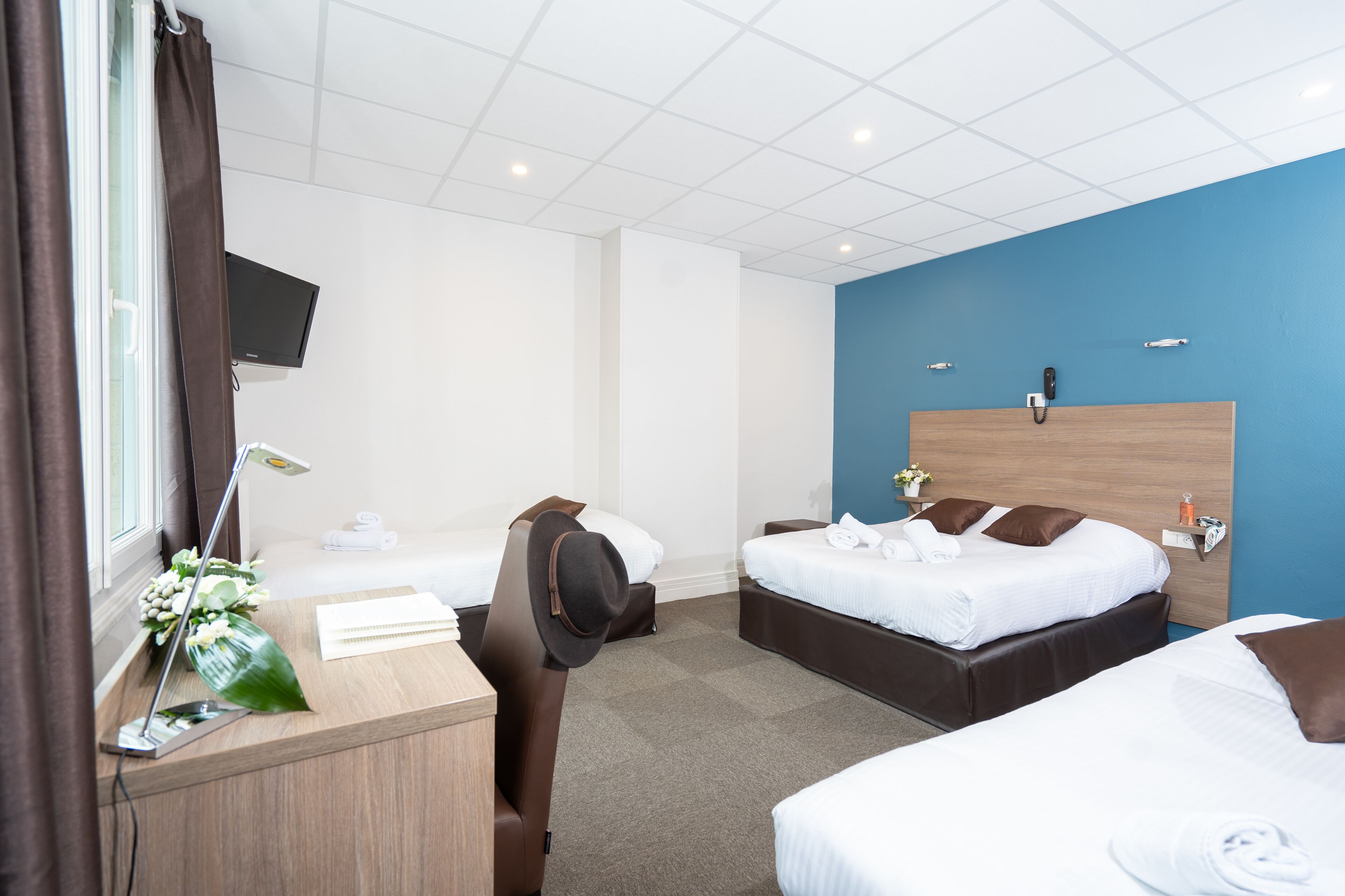 Quarto de hotel com duas camas, uma escrivaninha, uma cadeira, uma televisão e uma parede azul decorativa.