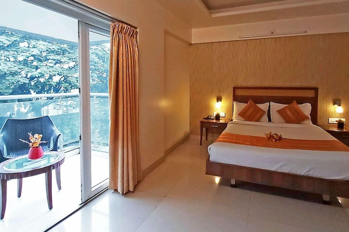 Quarto de hotel com cama, mesas de cabeceira, uma pequena mesa com cadeira e uma janela grande com cortinas.