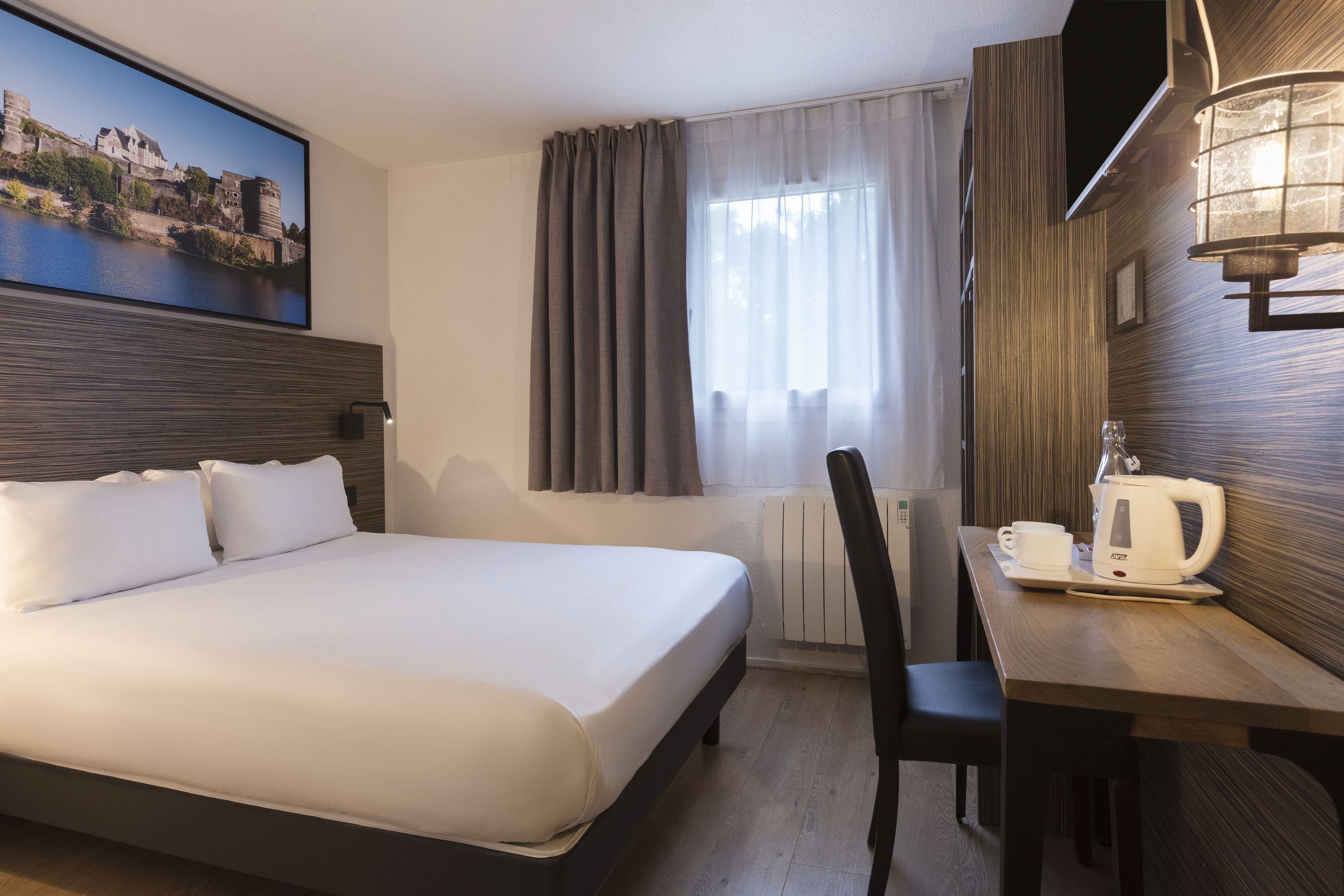 Quarto de hotel com cama, escrivaninha com chaleira, cadeira, janela com cortinas e um quadro na parede.