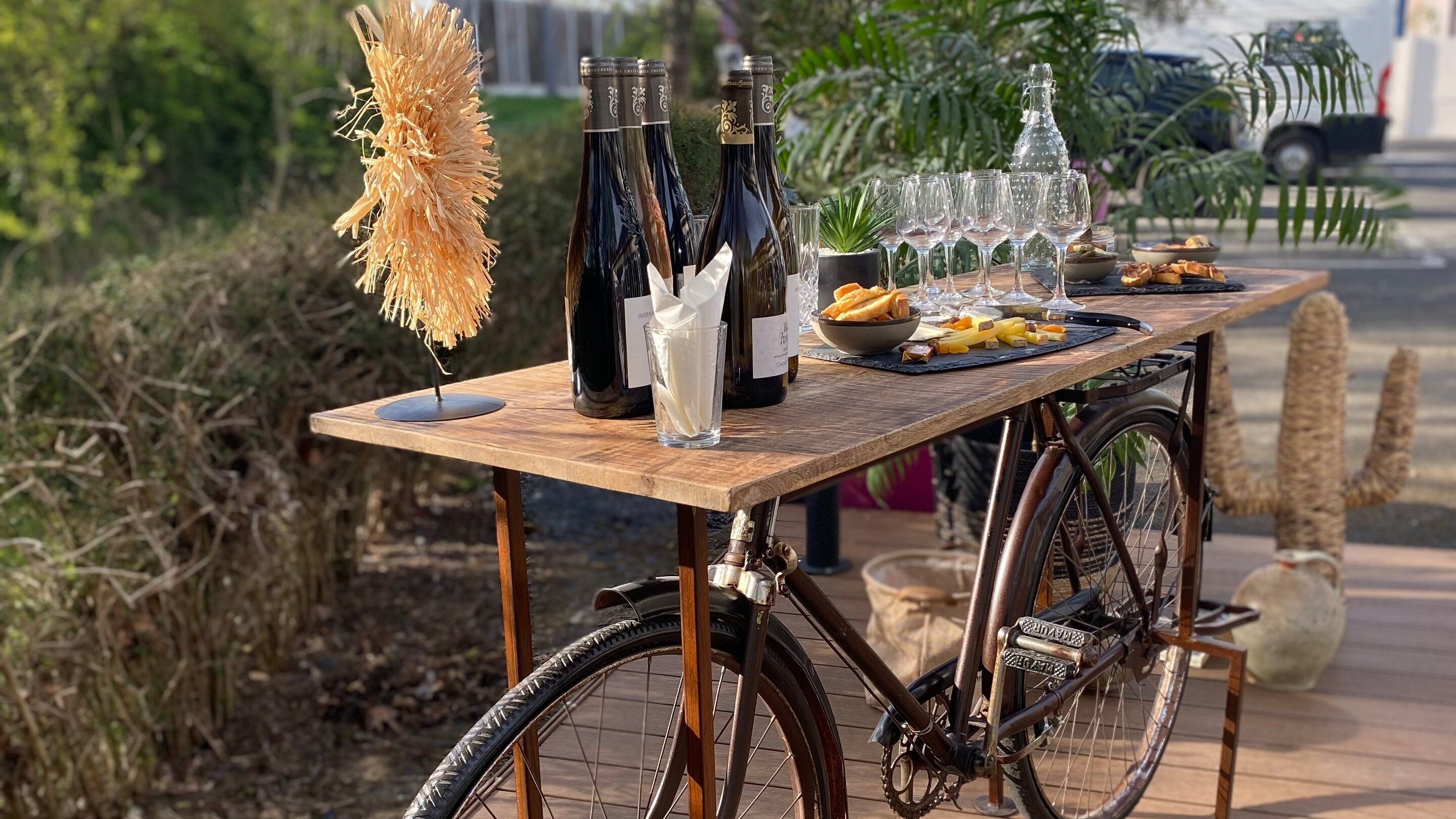 Carrinho de bicicleta com garrafas de vinho, copos e petiscos sobre uma mesa de madeira ao ar livre.