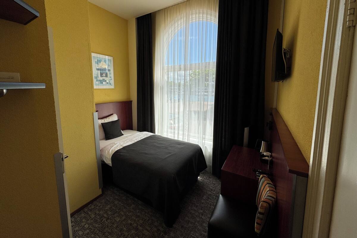 Quarto de hotel com cama, escrivaninha, cadeira, televisão e janela com cortinas.