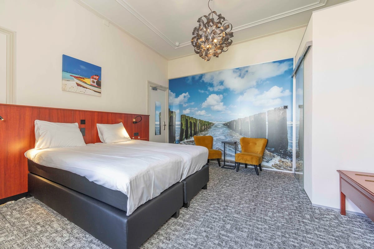 Quarto de hotel com cama, escrivaninha, duas cadeiras, um quadro e um mural.