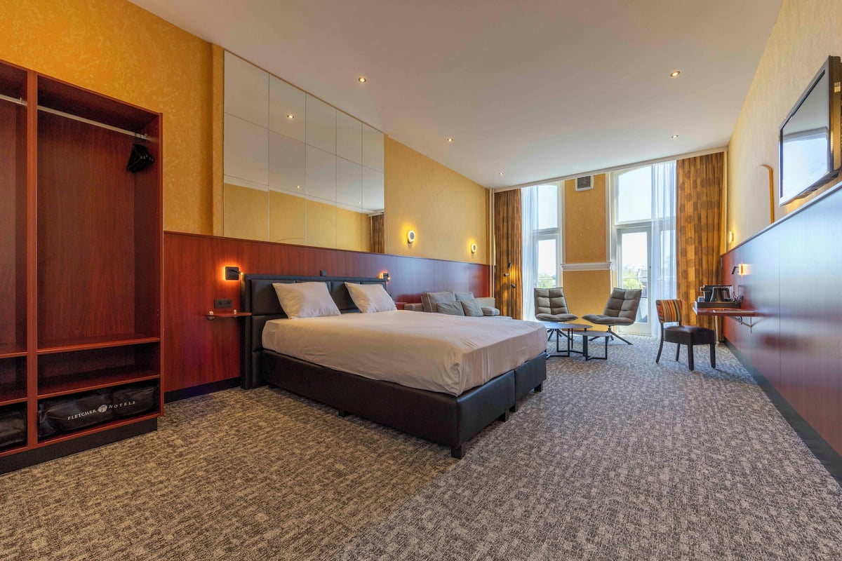 Quarto de hotel com cama grande, área de estar com cadeiras, escrivaninha e guarda-roupa.