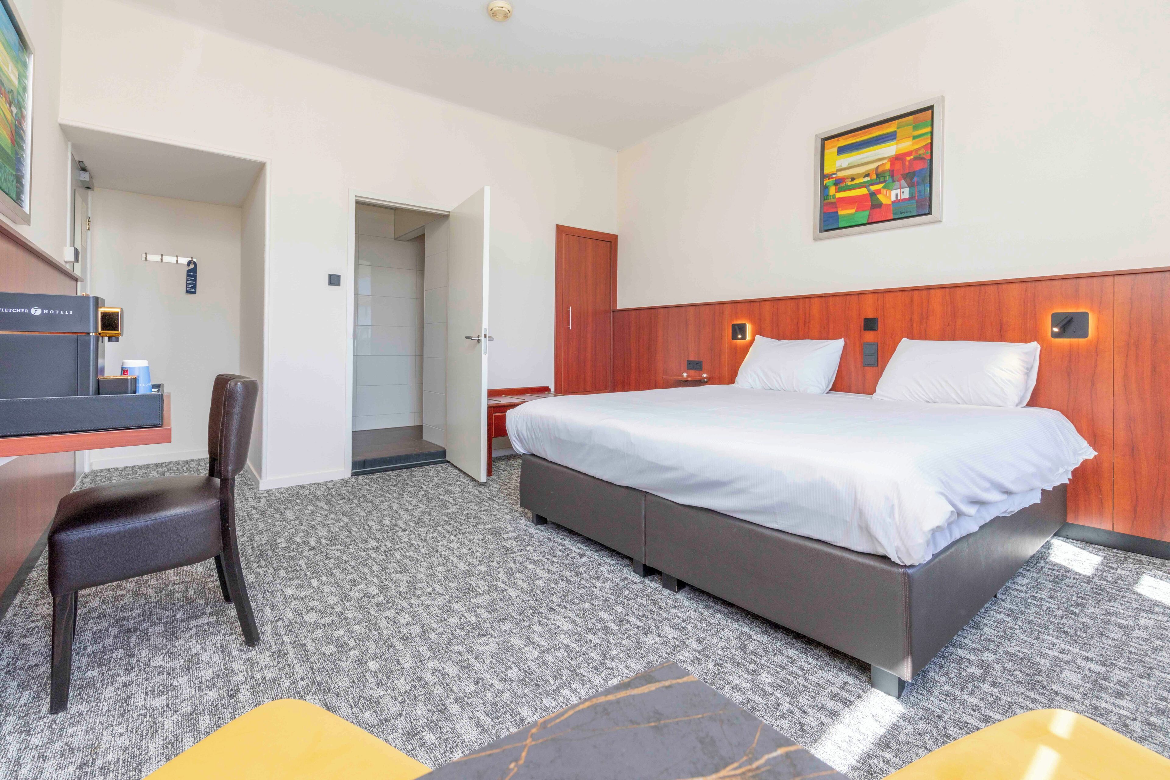 Quarto de hotel com cama, escrivaninha, cadeira e um quadro na parede.