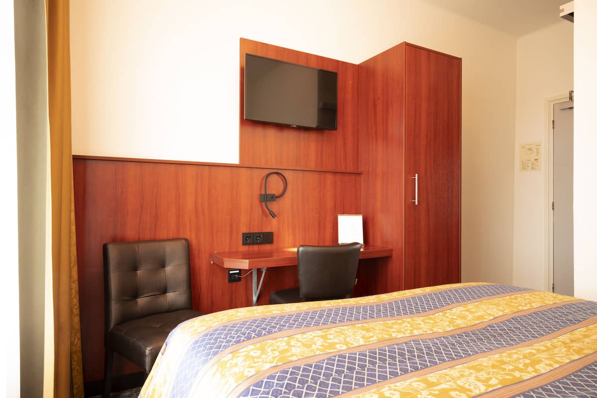 Quarto de hotel com cama, escrivaninha, cadeira, televisão e guarda-roupa.