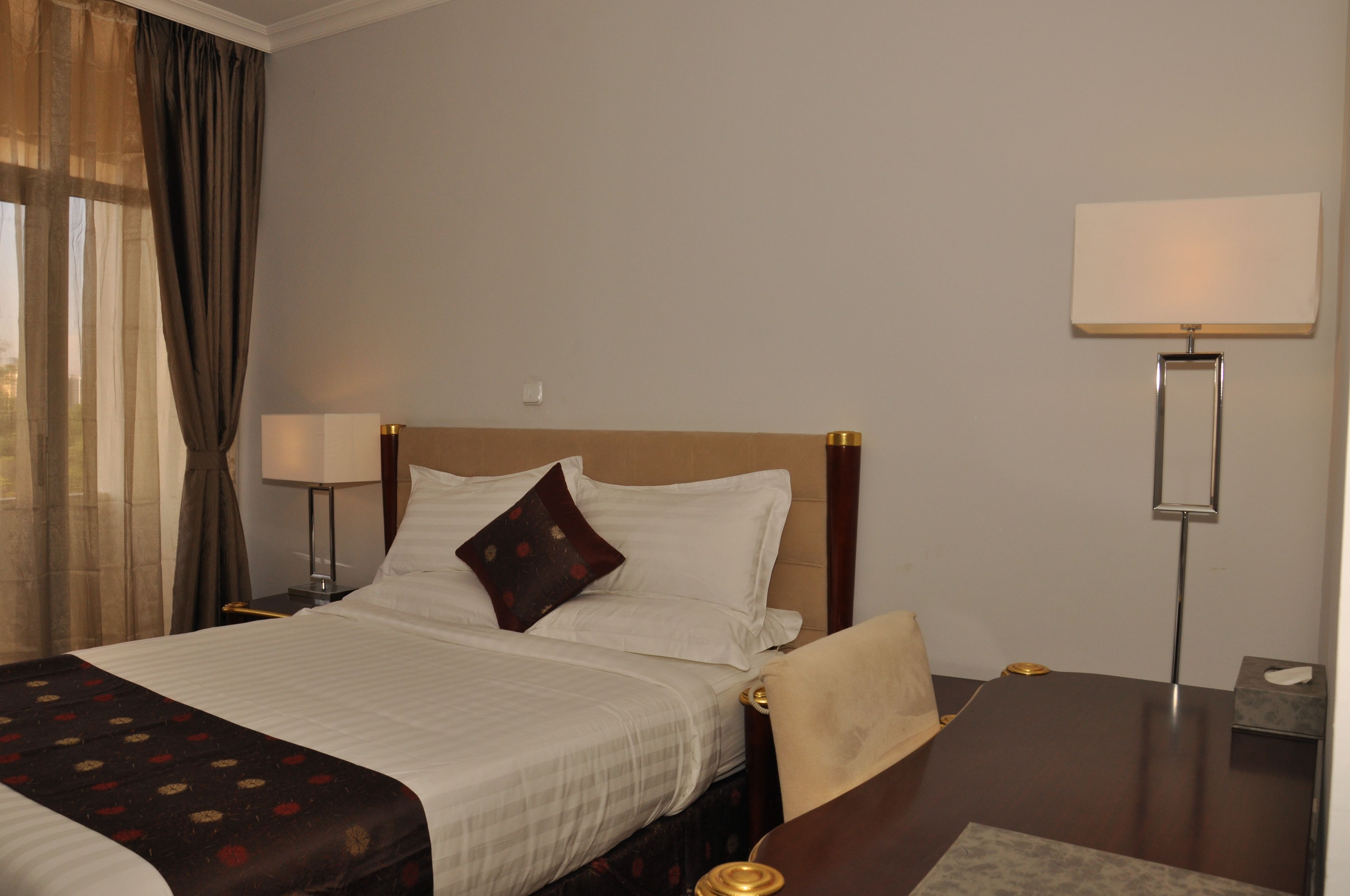 Quarto de hotel com uma cama bem arrumada, uma mesa de cabeceira com abajur, uma cadeira e uma escrivaninha com luminária.