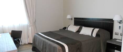 Quarto de hotel com uma cama grande, uma escrivaninha, uma cadeira, uma luminária e uma mesa de cabeceira.