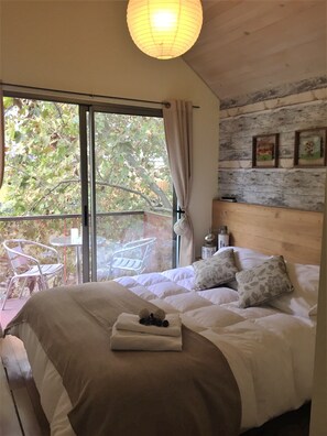 Quarto com cama, janela com vista para árvores, mesa com cadeiras do lado de fora e parede de madeira com quadros.