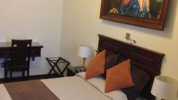 Quarto de hotel com cama, escrivaninha, cadeira, abajur e um quadro na parede.