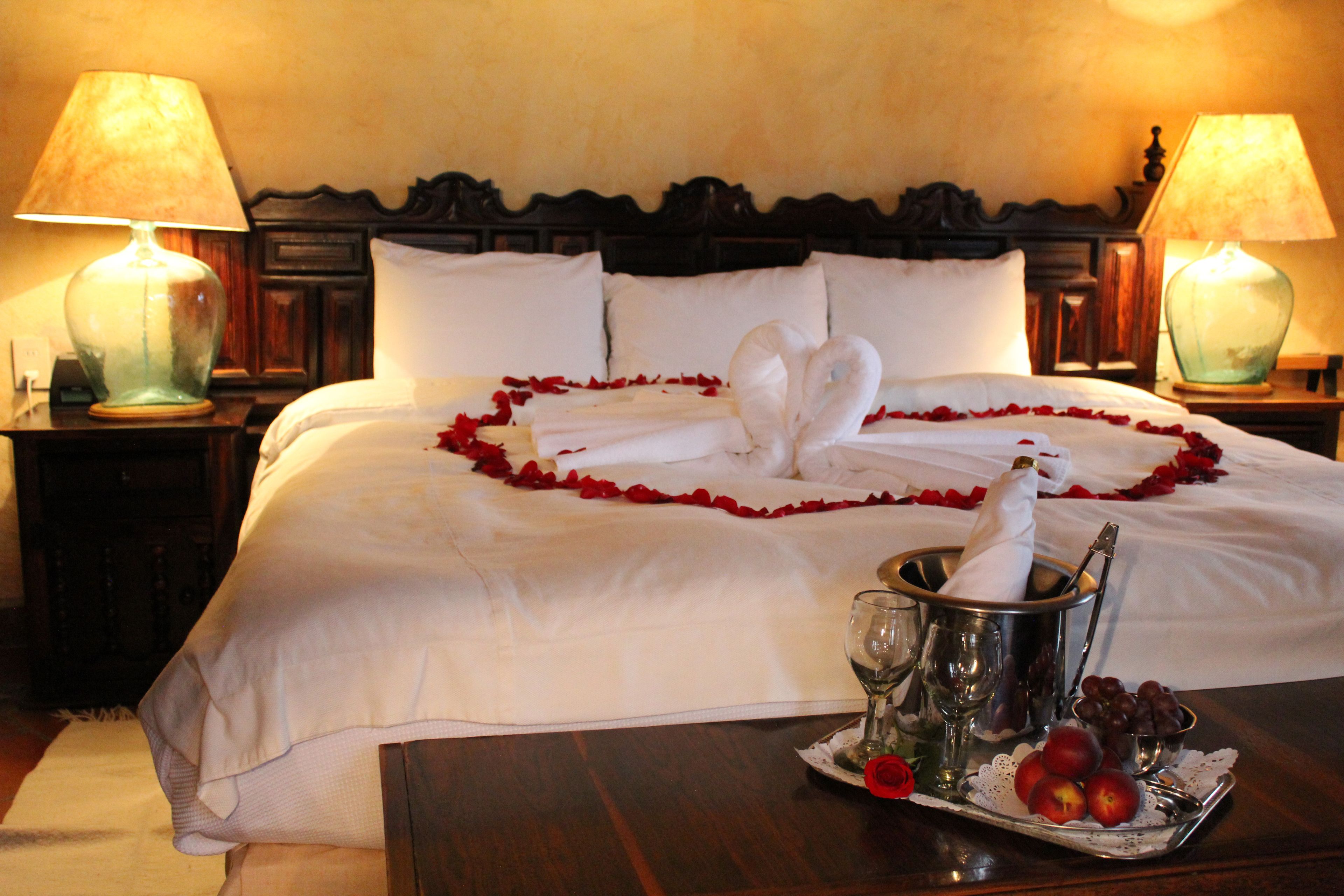 Quarto de hotel com cama arrumada, duas luminárias de cabeceira, uma bandeja com frutas e uma garrafa de champanhe, e um arranjo decorativo de pétalas de rosa na cama.