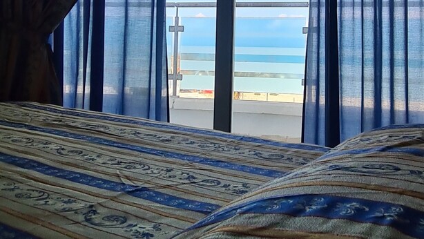 Quarto com cama, vista para a praia e varanda com cortinas azuis.