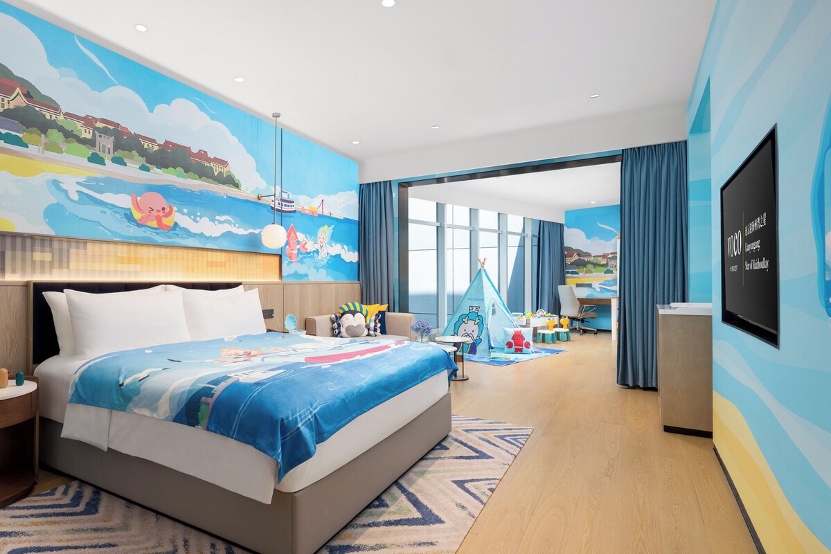 Quarto de hotel com uma cama grande, uma mesa, uma cadeira, uma televisão e um mural na parede.