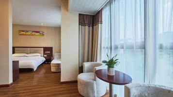 Quarto de hotel com uma cama, uma mesa redonda com uma planta, uma cadeira e uma janela grande com cortinas.