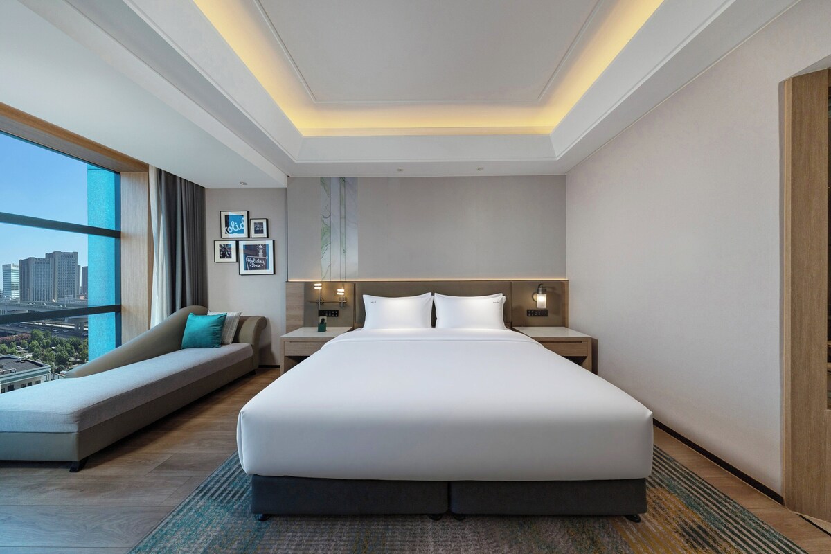 Quarto de hotel moderno com uma cama grande, um sofá, uma mesinha de cabeceira e vista para a cidade.