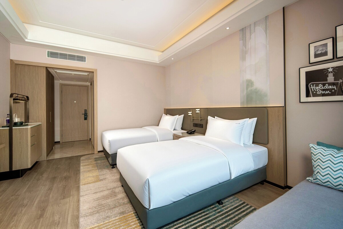 Quarto de hotel com duas camas, uma escrivaninha, um espelho e uma parede com quadros.