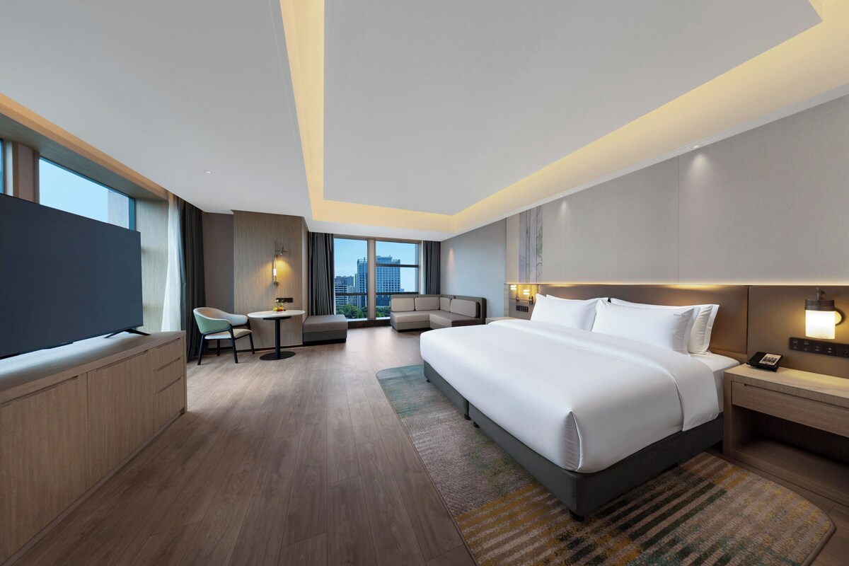 Quarto de hotel moderno com uma cama grande, área de estar com sofá e poltronas, televisão de tela plana e uma janela ampla com vista para a cidade.