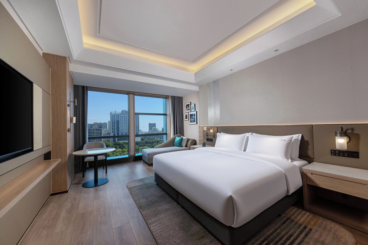 Quarto de hotel moderno com uma cama grande, uma mesa de trabalho, uma televisão e vista para a cidade.