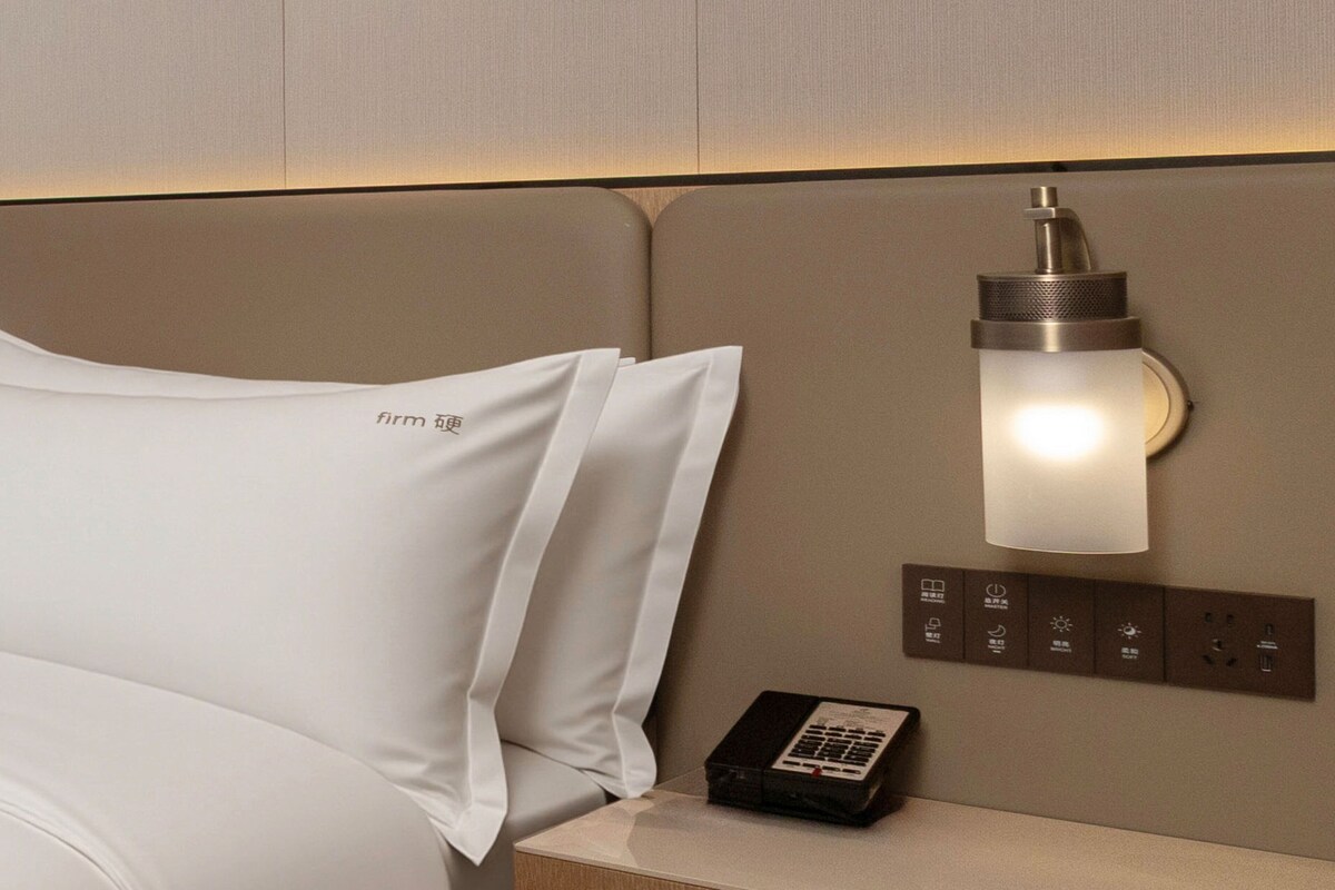 Quarto de hotel com cama, mesa de cabeceira com abajur e painel de controle com vários ícones.