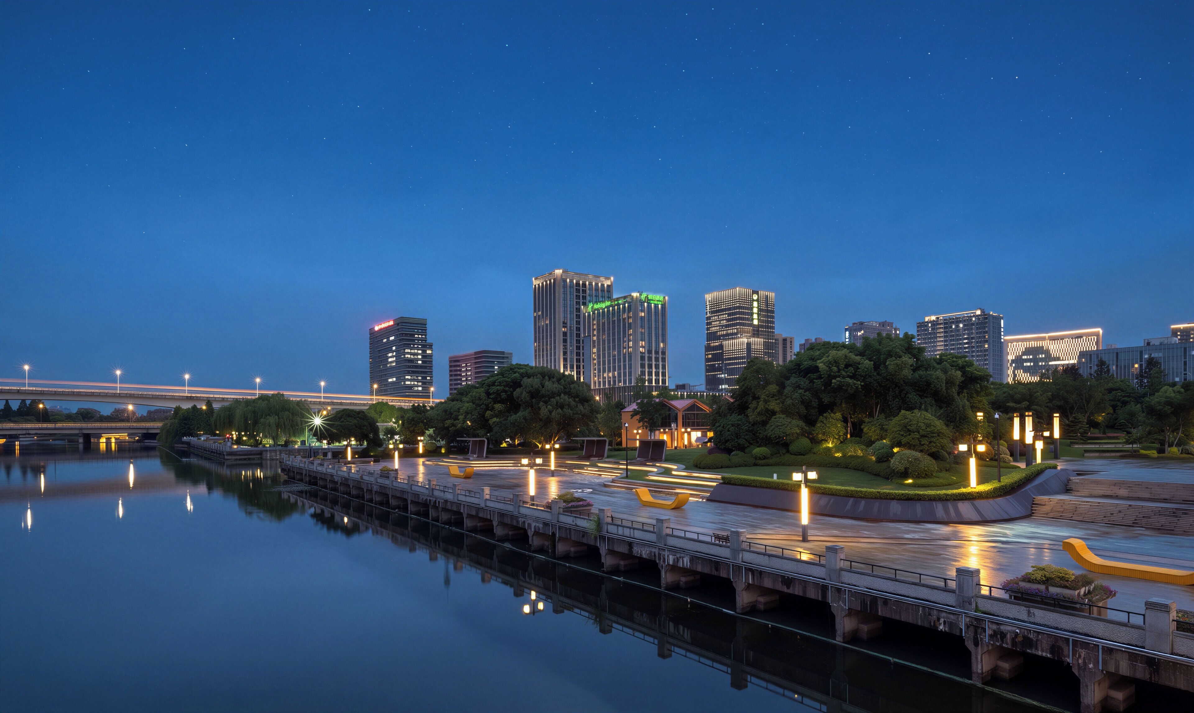 Paisagem urbana ao entardecer, com edifícios iluminados, um rio calmo e uma ponte.