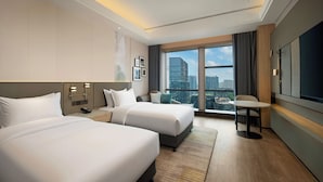 Quarto de hotel com duas camas, uma escrivaninha, uma cadeira, televis ão e uma janela grande com vista para a cidade.