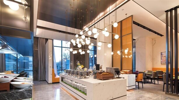 Um hall de hotel moderno com um balcão central para o café da manhã, janelas amplas e luminárias contemporâneas.