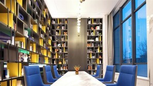 Uma biblioteca moderna com uma mesa comprida e cadeiras azuis, rodeada por estantes altas com vários livros e objetos decorativos.