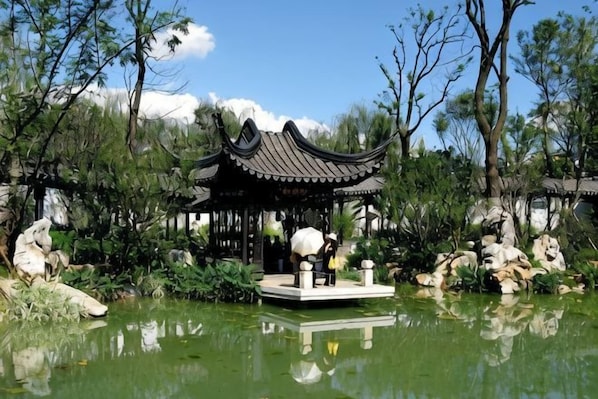 Um pavilhão tradicional com telhado de azulejos, rodeado por um lago e pedras, com árvores ao fundo.
