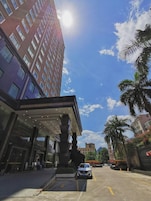 Entrada de hotel com galeria coberta, estacionamento e um edifício alto ao fundo.