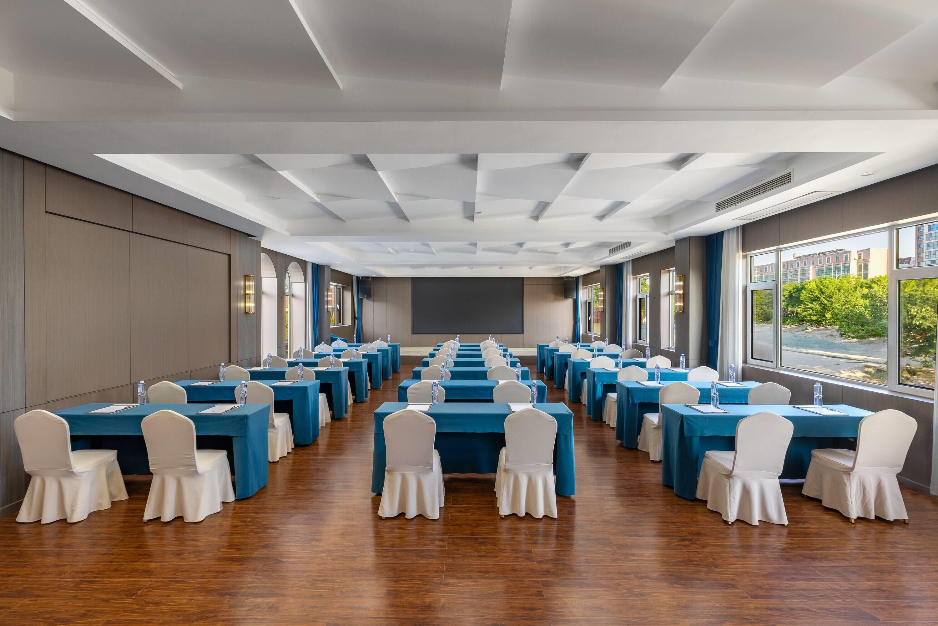 Sala de conferências com fileiras de mesas e cadeiras, amplas janelas e design moderno no teto.