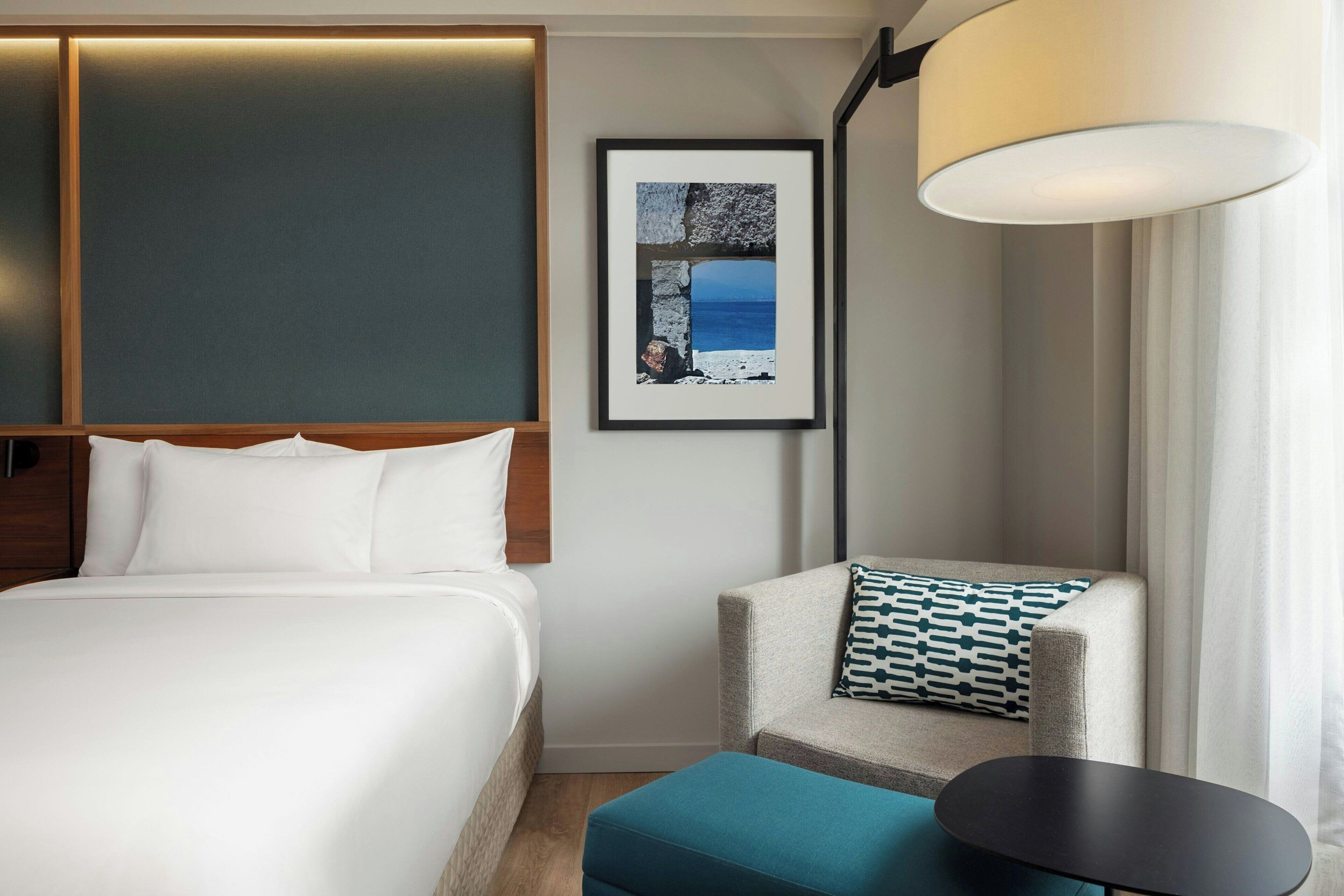 Quarto de hotel com uma cama, uma poltrona pequena, uma mesinha de cabeceira e um quadro na parede.