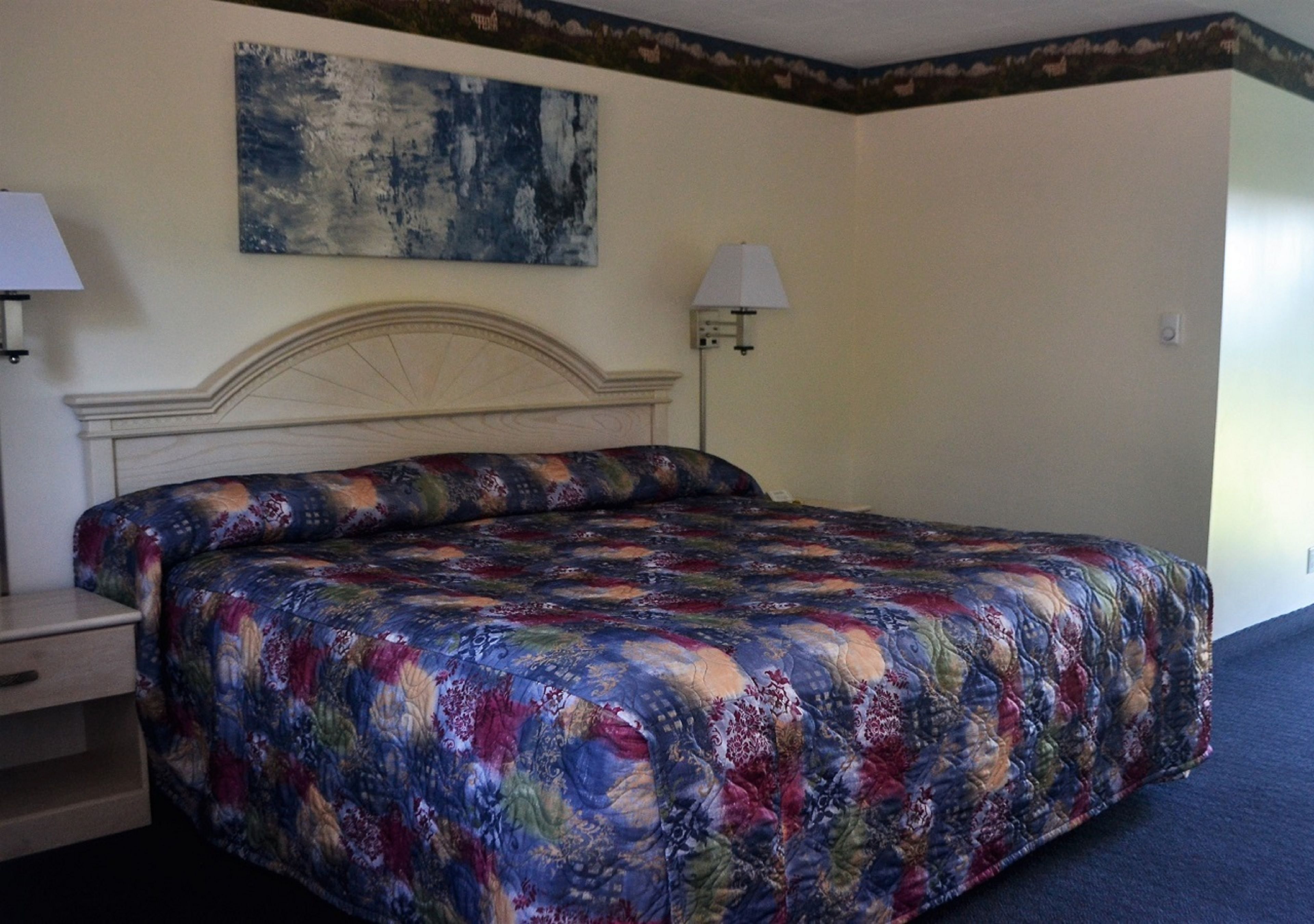 Quarto com cama, duas luminárias de cabeceira, mesa de apoio e um quadro na parede.