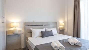 Cama bem arrumada com lençóis brancos e cinzas, duas luminárias de cabeceira e toalhas enroladas sobre a cama.