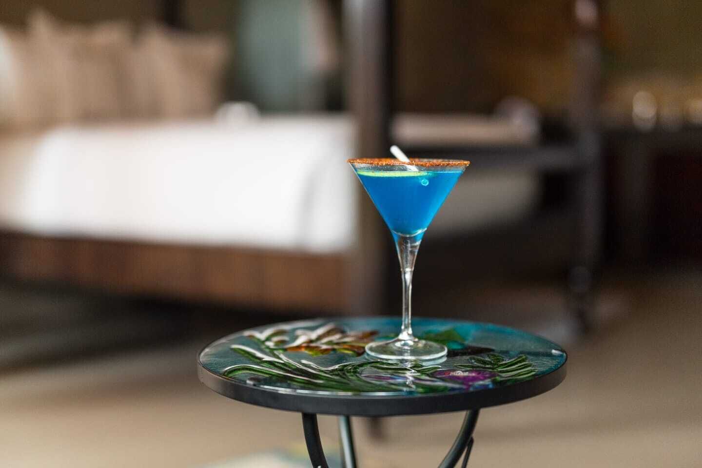 Um cocktail azul num copo de martini sobre uma mesa redonda com tampo de vidro, num quarto com uma cama ao fundo.