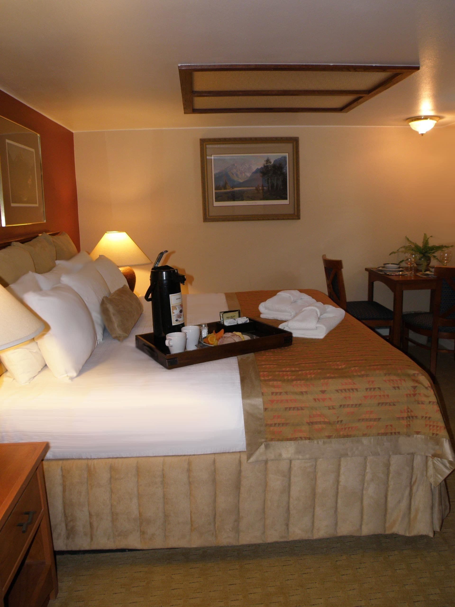 Quarto de hotel com uma cama, uma mesinha de cabeceira com luminária, um tabuleiro com itens de café da manhã, um quadro na parede e uma pequena mesa com uma planta.