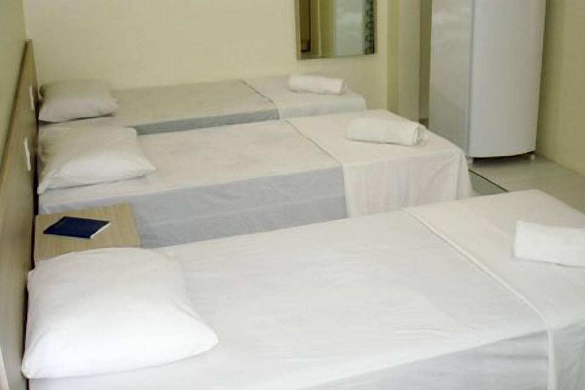 Quarto de hotel com três camas de solteiro, cada uma com travesseiro e cobertor. Há uma mesinha com um livro e uma geladeira no canto.