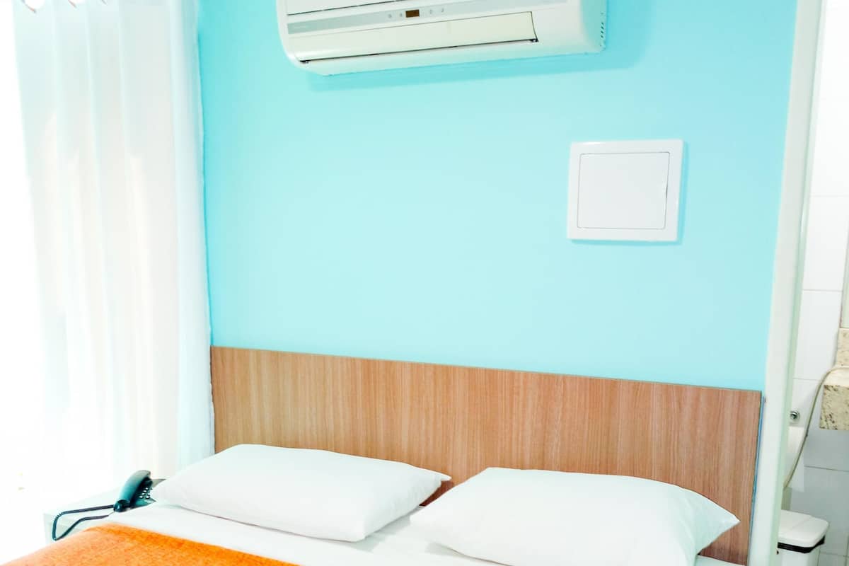 Quarto de hotel com cama, unidade de ar condicionado, travesseiro branco e cobertor branco.