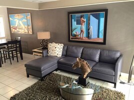 Uma sala de estar com um sofá seccional, uma mesa de jantar com cadeiras, uma mesa de centro com um objeto decorativo e duas pinturas na parede.