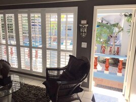 Quarto com uma cadeira preta, uma mesa de vidro e vista através de uma porta de correr para área externa com plantas e uma piscina.